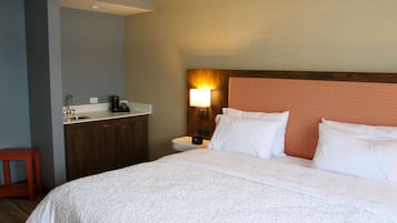 Quarto de hotel com uma cama grande, uma mesa de cabeceira com abajur, uma pequena escrivaninha com cafeteira e um armário de madeira.