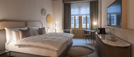 Quarto de hotel moderno com uma cama grande, uma escrivaninha, uma cadeira e vista para o exterior.
