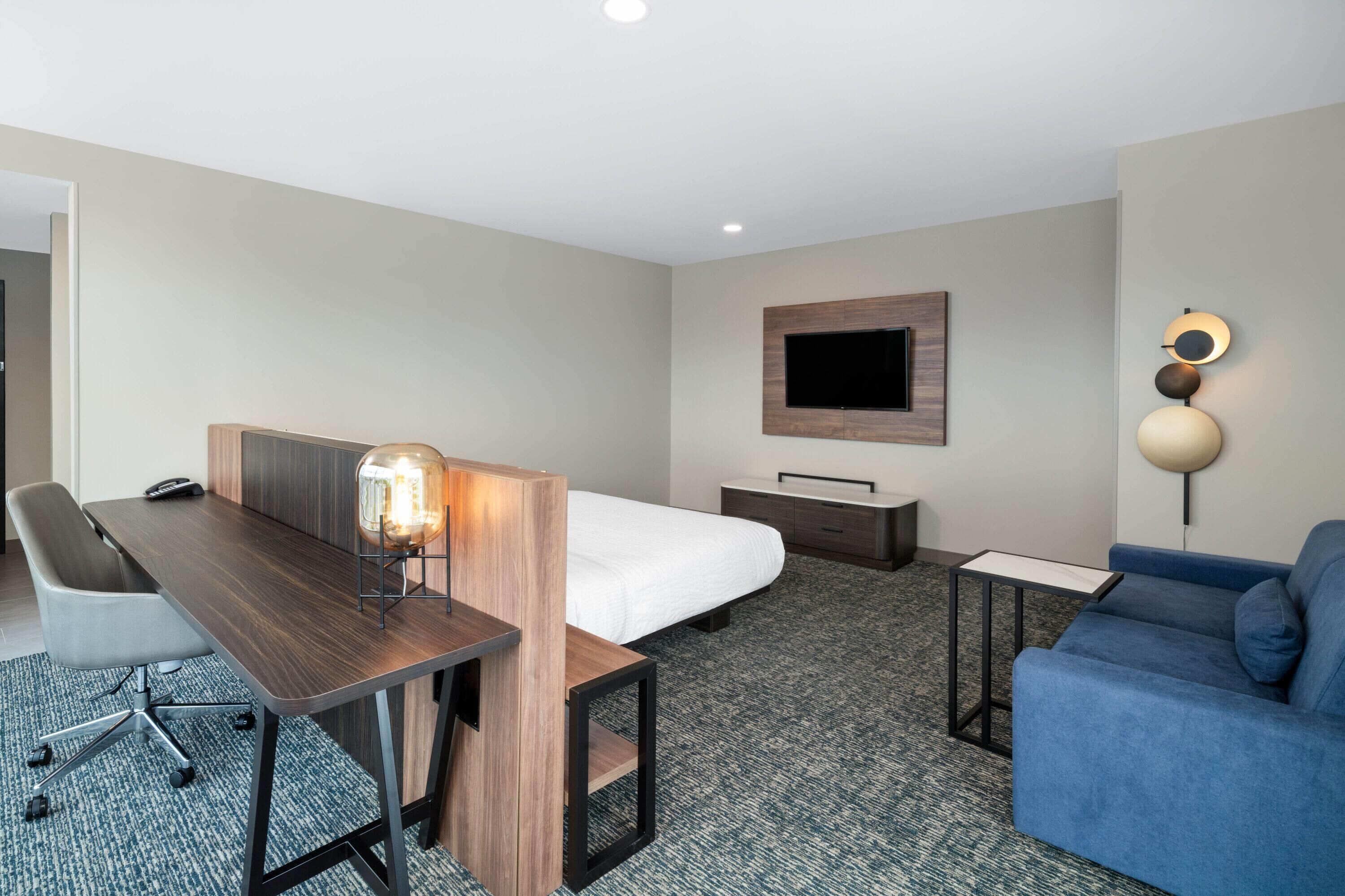 Quarto de hotel com escrivaninha de madeira, sofá azul, cama, televisão fixada na parede e uma mesinha.