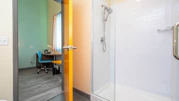 Casa de banho de hotel com chuveiro em cabine de vidro, sanita e secretária com cadeira.