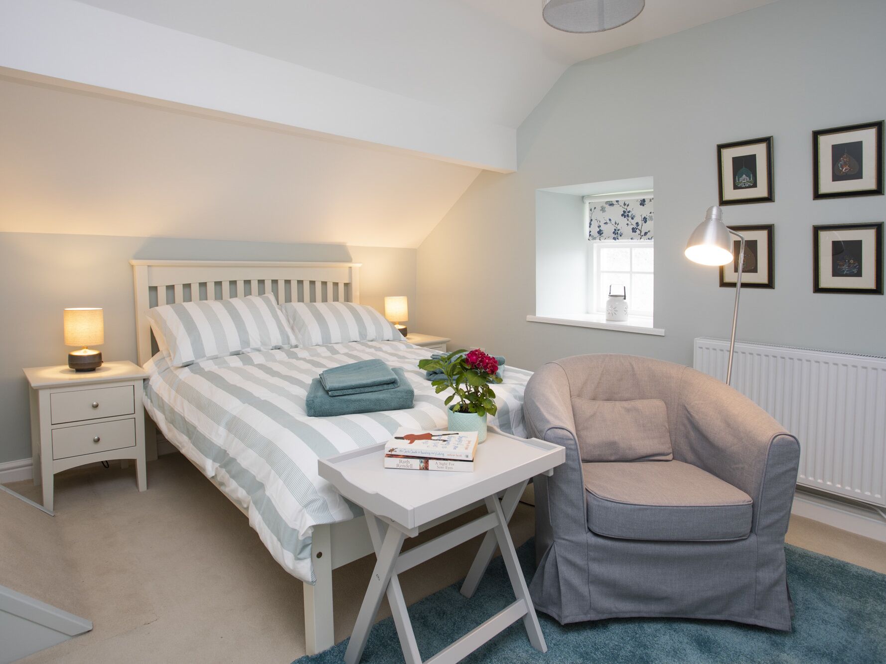 Quarto com cama, mesas de cabeceira, uma cadeira, janela e quadros nas paredes.