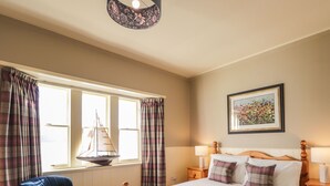 Quarto com cama de madeira, uma cadeira, mesa de cabeceira, luminária, janela com cortinas, modelo de veleiro e um quadro na parede.