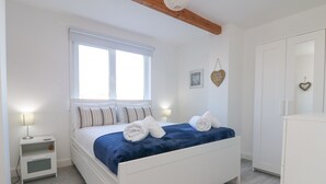 Quarto com cama branca, janela, mesa de cabeceira, guarda-roupa e uma decoração em formato de coração.