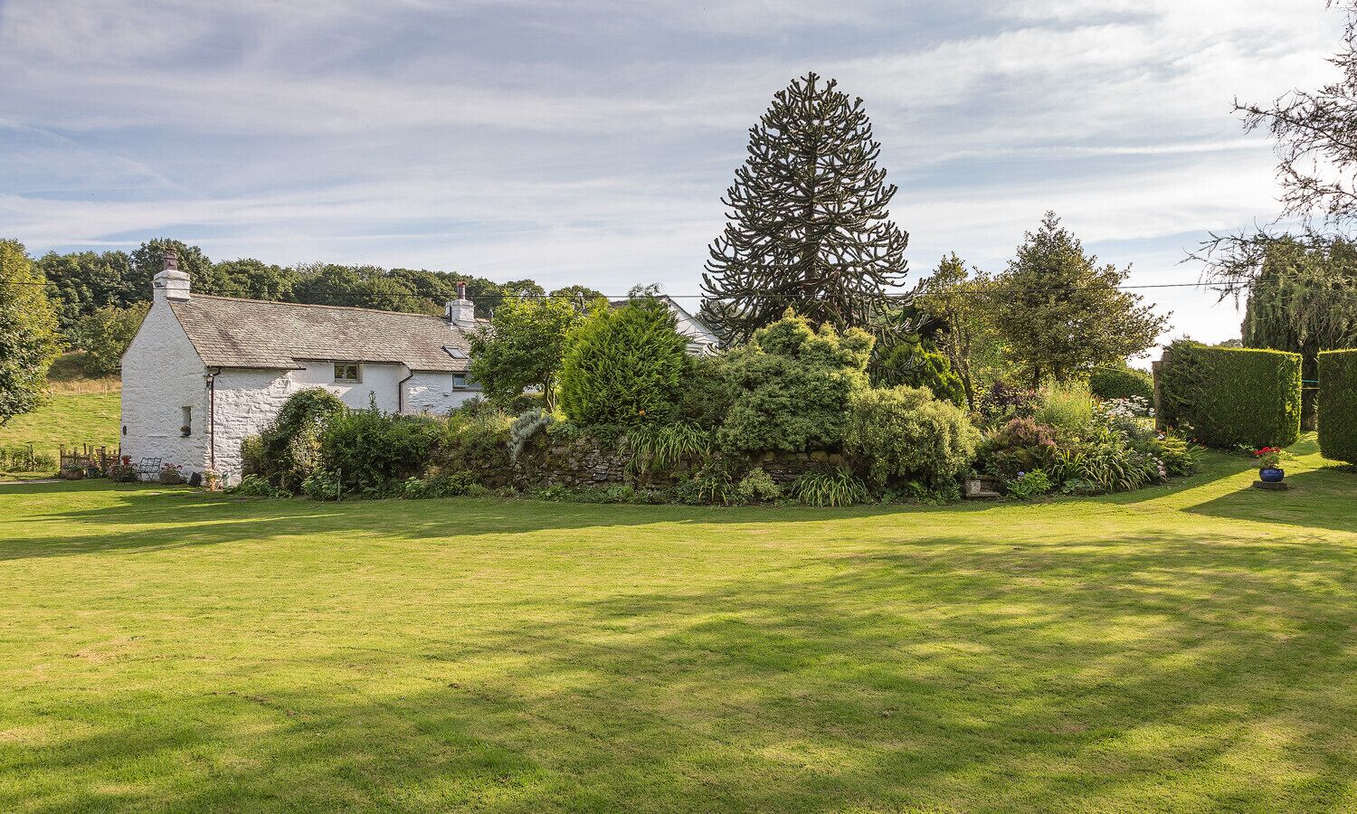 Uma casa de pedra branca com telhado inclinado, cercada por um gramado bem cuidado e vegetação densa.