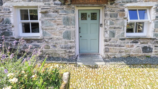 Uma casa de pedra com porta verde, duas janelas pequenas e uma placa acima da porta com a inscrição "Yew Tree Cottage".