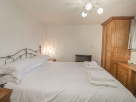 Uma cama bem arrumada com roupa de cama branca, uma mesinha de cabeceira de madeira com uma luminária, um radiador e um guarda-roupa de madeira.