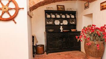 Uma cozinha rústica com piso de tijolos, um leme de madeira, um armário preto com pratos brancos e uma cesta de vime com flores vermelhas.
