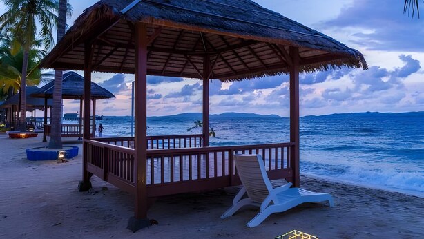 Um gazebo à beira-mar com telhado de palha, uma espreguiçadeira e um pequeno barco.