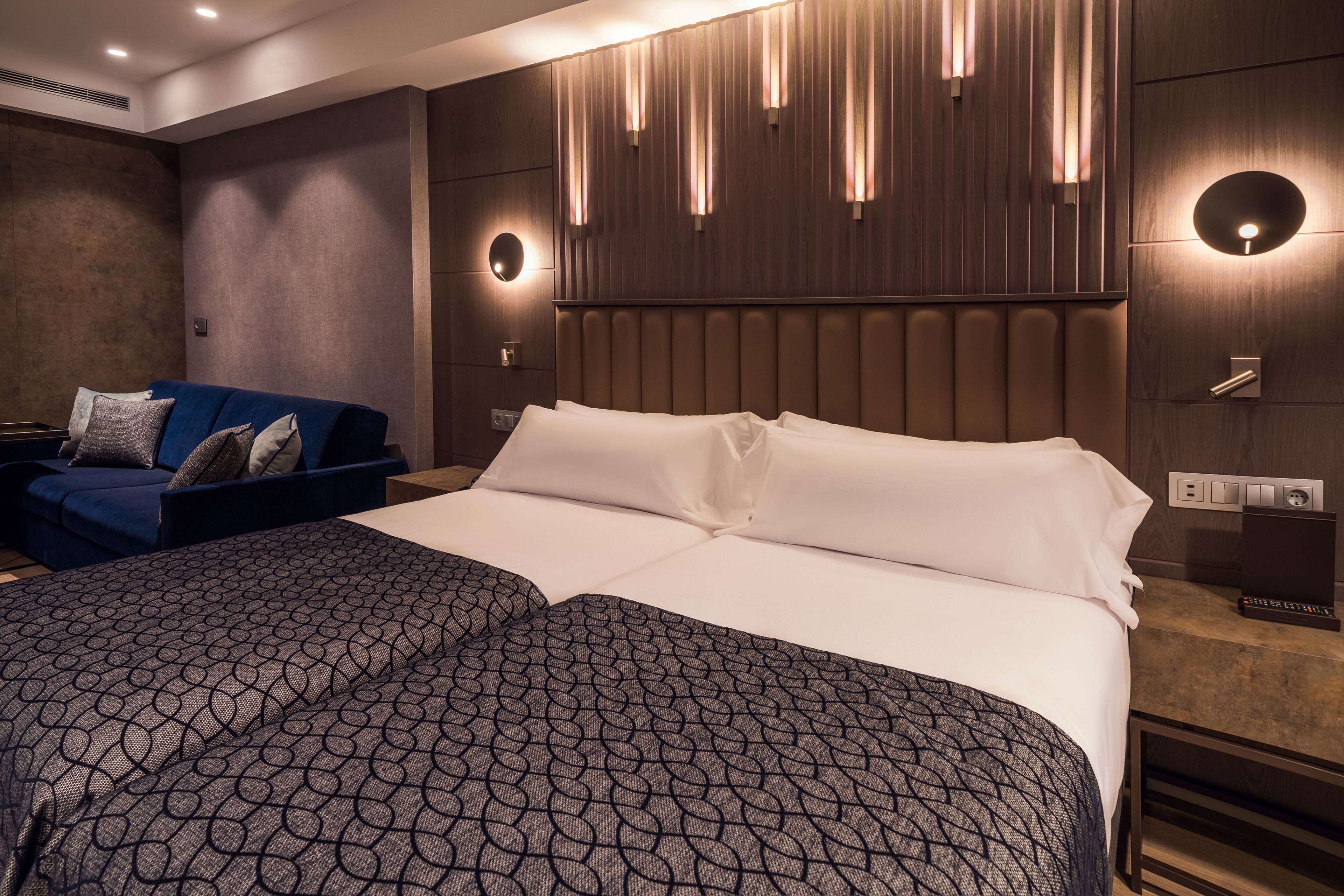 Quarto de hotel com uma cama grande, um sofá azul e uma luminária fixada na parede.