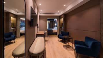 Quarto de hotel moderno com piso de madeira, um espelho grande, televisão de tela plana, sofá, mesa pequena e uma cama com cabeceira azul.