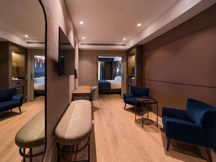 Quarto de hotel moderno com piso de madeira, um espelho grande, televisão de tela plana, sofá, mesa pequena e uma cama com cabeceira azul.
