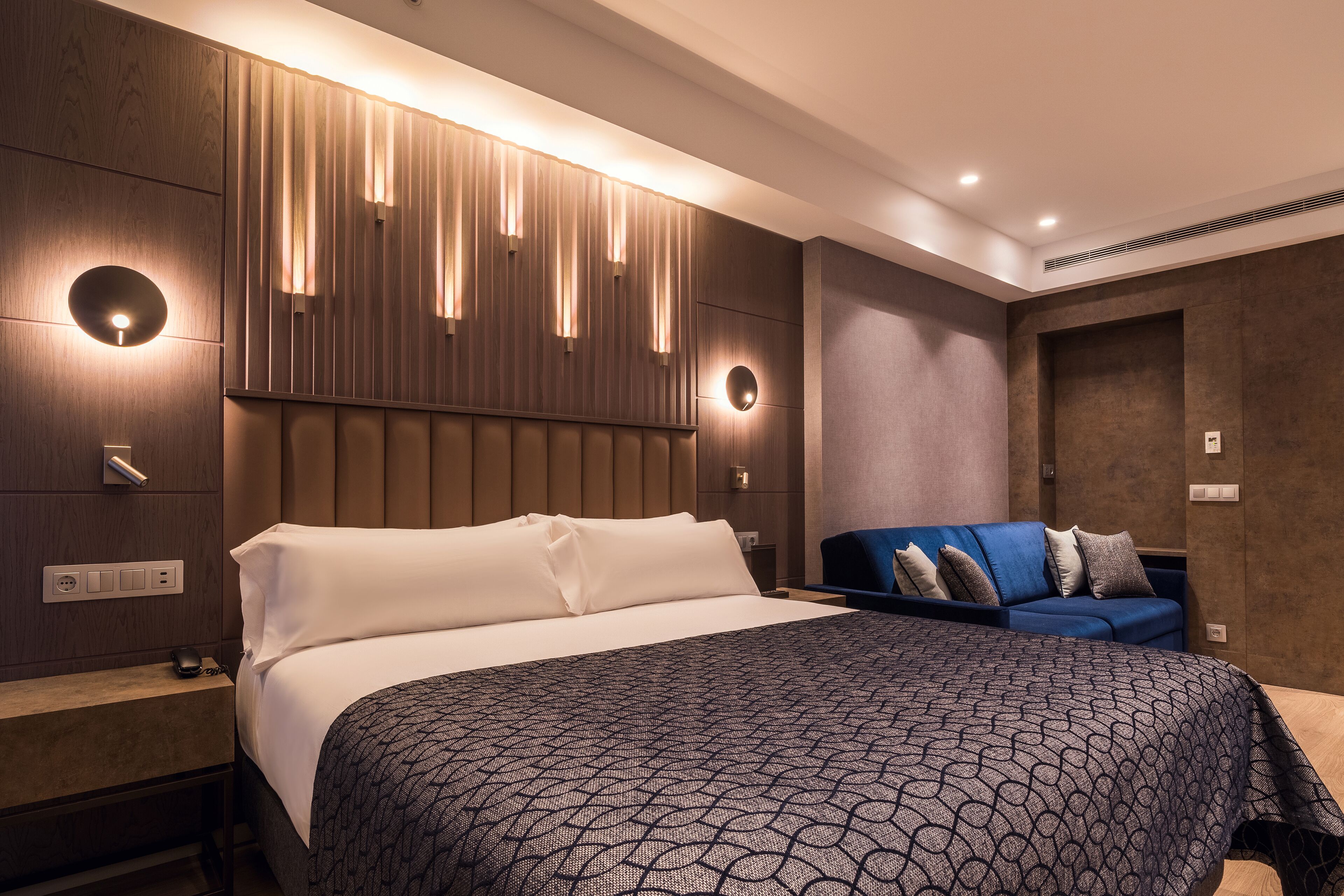 Quarto de hotel moderno com uma cama grande, um sofá azul e um candeeiro fixado na parede.
