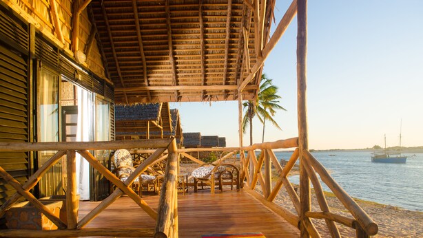 Um deck de madeira com cadeiras e uma mesa, com vista para uma praia com barcos e palmeiras.