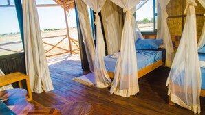 Quarto com cama sob dossel, piso de madeira e vista para a praia.