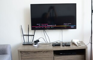 Uma televisão fixada na parede com um centro de entretenimento de madeira abaixo. A TV exibe um canal de música com o título "Life Catcher" e o artista "Khamis".