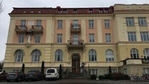 Um edifício com vários andares, uma entrada central, varandas e uma área de estacionamento com carros e bicicletas.