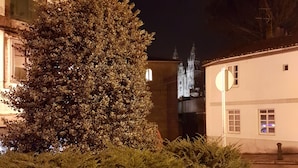 Cena noturna de rua com um edifício em primeiro plano, apresentando uma grande árvore, e uma igreja ao fundo, com torres iluminadas.