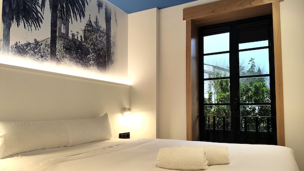 Quarto de hotel com uma cama grande, uma mesa de cabeceira, uma luminária na parede e uma janela com vista para o verde.