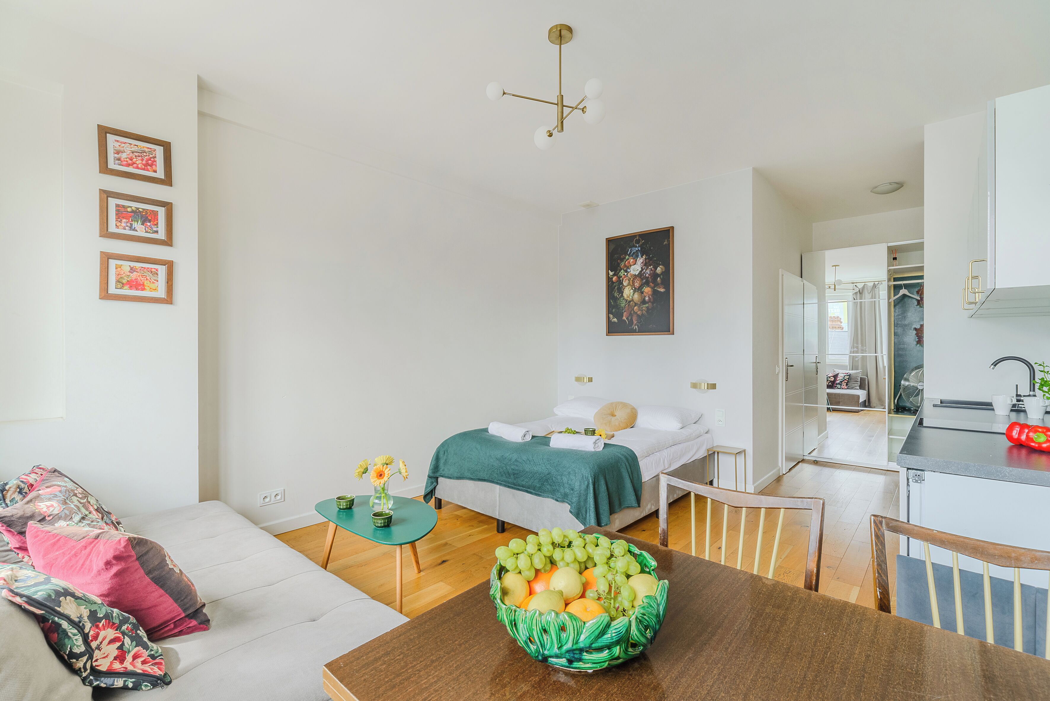 Quarto moderno e bem iluminado, com uma cama, uma mesa de jantar com uma cesta de frutas, um sofá com almofadas florais e uma cozinha visível através de uma porta aberta.