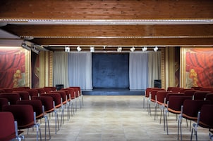 Uma sala de conferências com fileiras de cadeiras vermelhas, um palco com cortinas e teto de madeira.