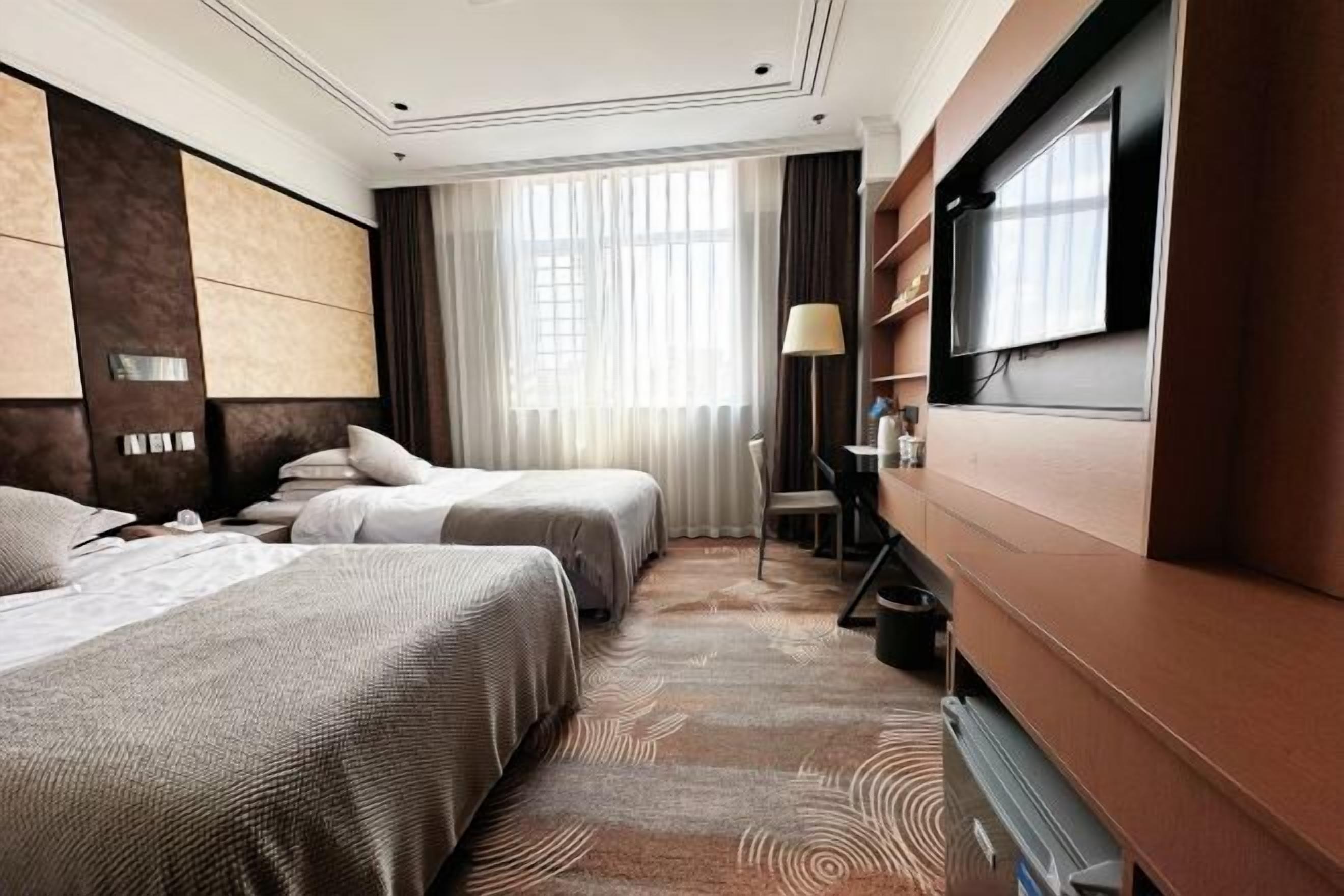 Quarto de hotel com duas camas, televisão, escrivaninha e uma janela grande com cortinas.