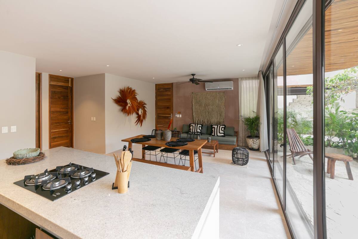 Uma cozinha moderna com fogão a gás, mesa de jantar de madeira e cadeiras. Há uma sala de estar com sofá e uma janela ampla com vista para um jardim.