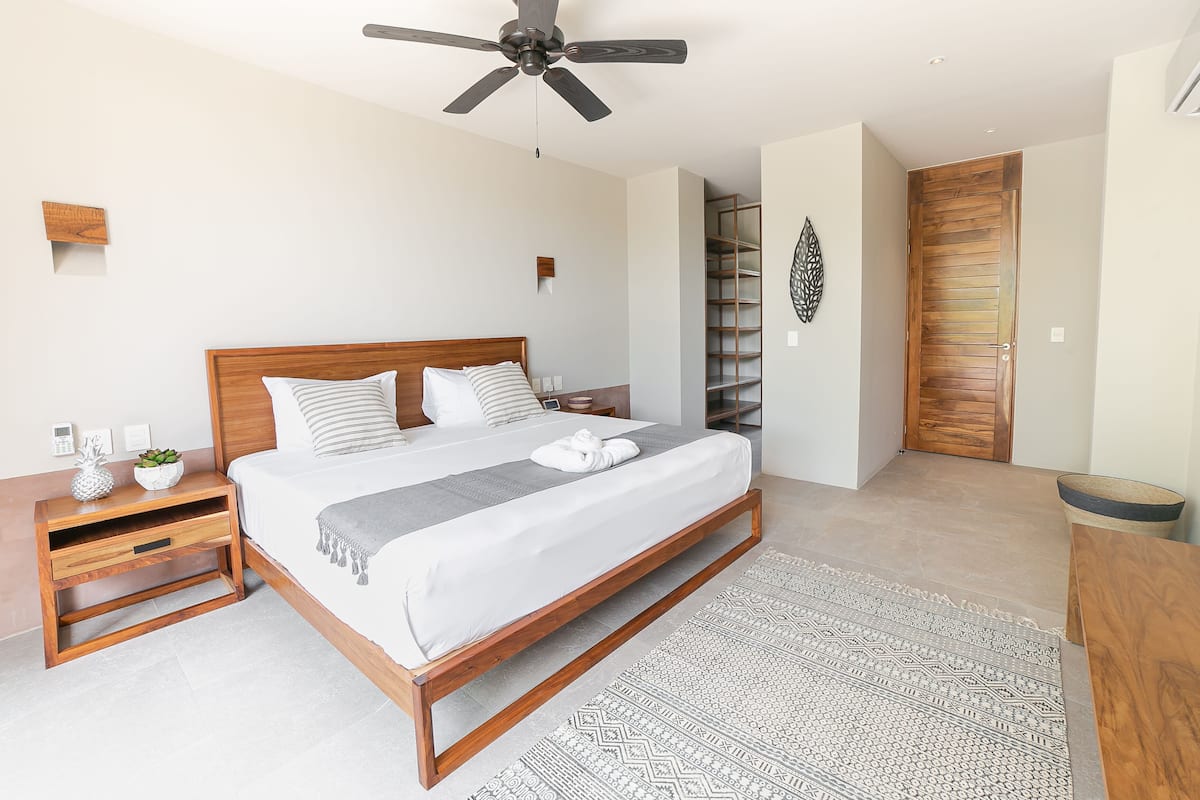 Quarto moderno com cama de madeira, ventilador de teto, mesa de cabeceira com uma planta e tapete estampado.