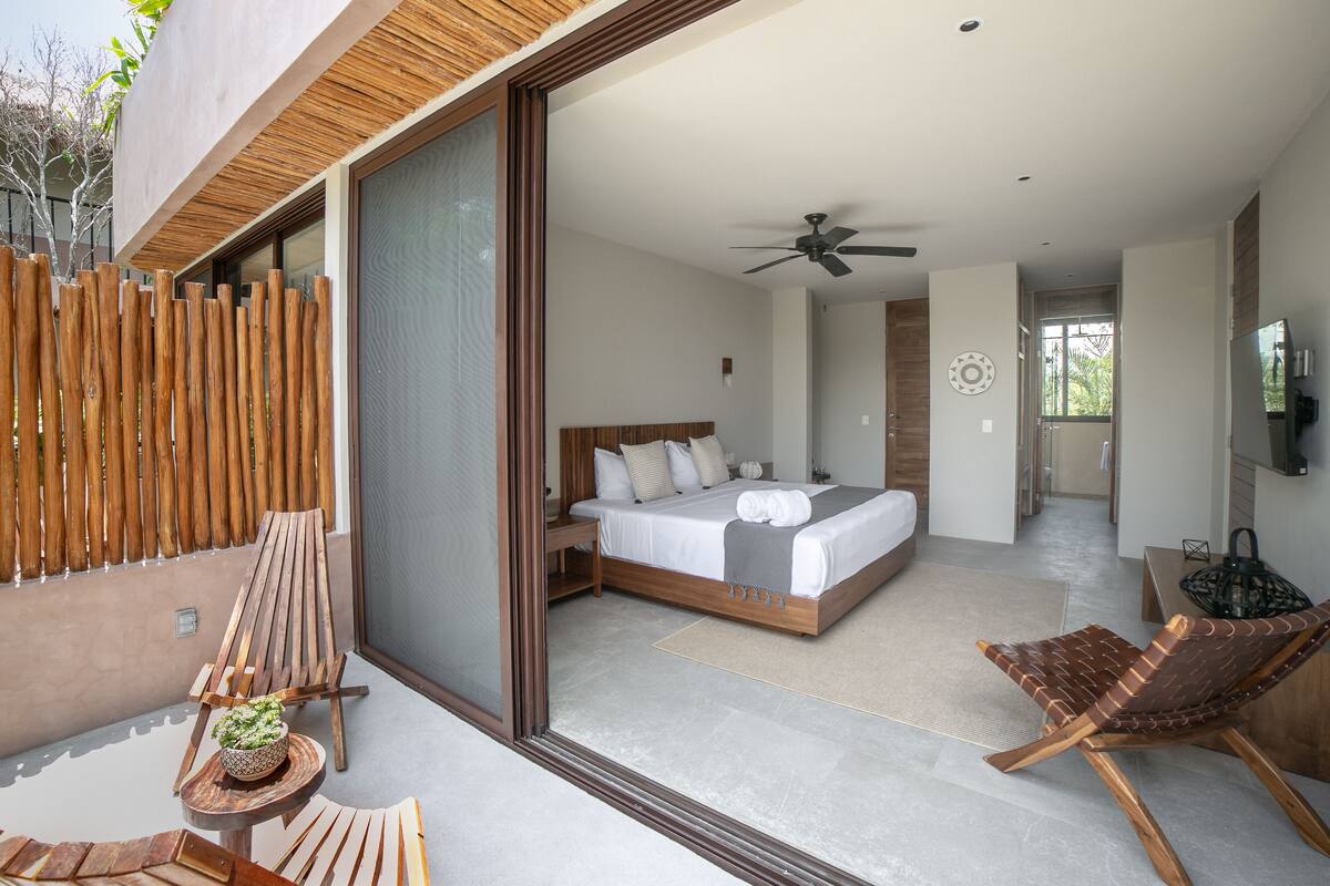 Um quarto moderno com uma cama grande, um ventilador de teto e vista para uma varanda por uma porta deslizante, onde há uma cadeira de madeira e uma mesa pequena.