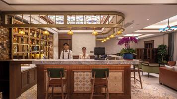 Uma área de bar com bancada de mármore, dois bancos altos e um bartender. Há prateleiras com garrafas atrás do balcão, uma grande planta em vaso e uma escadaria com um vitral ao fundo.