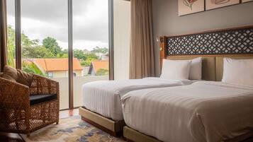 Quarto de hotel com duas camas, uma cadeira de vime, uma janela grande com vista para vegetação e edifícios, e obras de arte na parede.