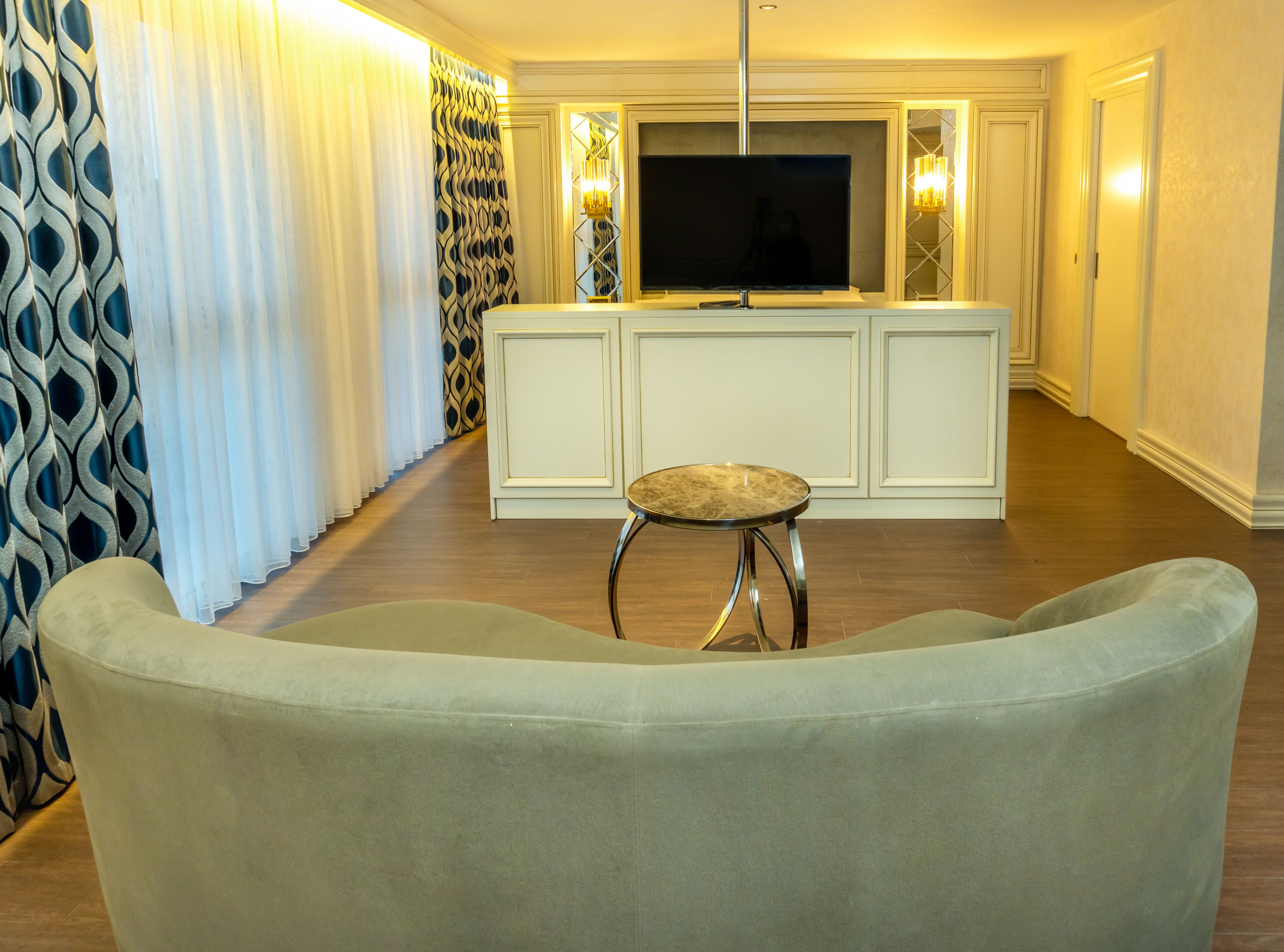 Uma sala de estar moderna com um sofá grande, uma mesa redonda pequena, uma TV de tela plana e cortinas estampadas.