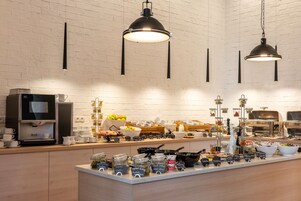 Buffet com diversos alimentos, acompanhamentos, máquina de café e luminárias pendentes.