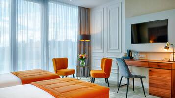 Quarto de hotel com cama, duas cadeiras, escrivaninha, televisão e abajur.