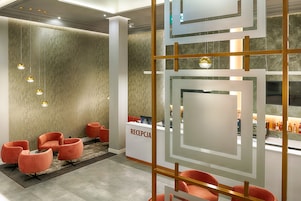 Uma área de recepção com design moderno, com um balcão de atendimento identificado com a palavra "RECEPTION" e algumas cadeiras laranja dispostas para hóspedes.