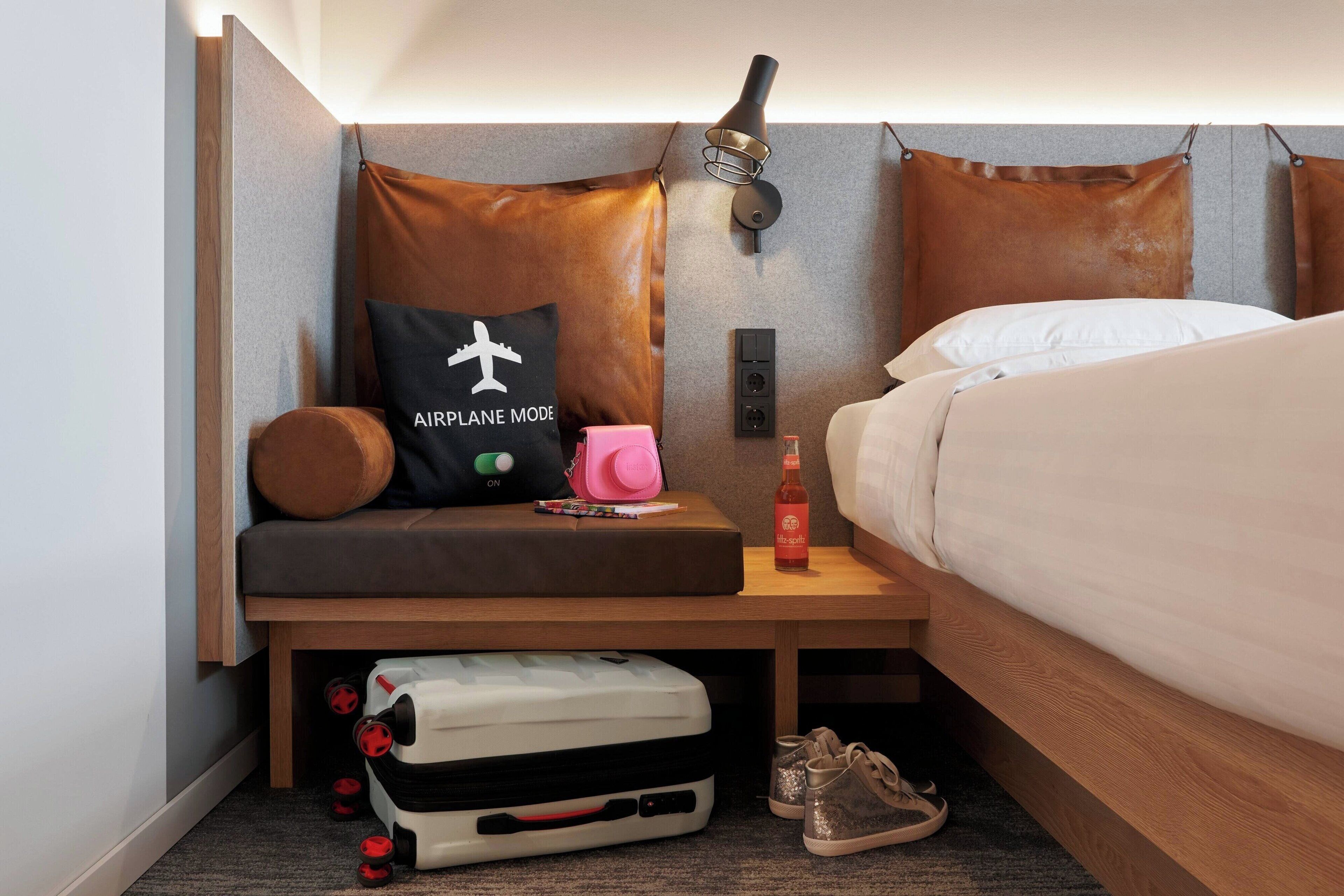 Quarto de hotel pequeno, com uma cama, mesa de cabeceira, um cantinho de estar com um travesseiro, luminária e mala.