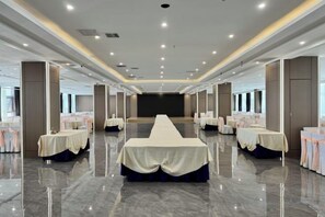 Uma ampla sala de banquetes com uma mesa comprida no centro, rodeada por mesas menores, e piso lustroso.