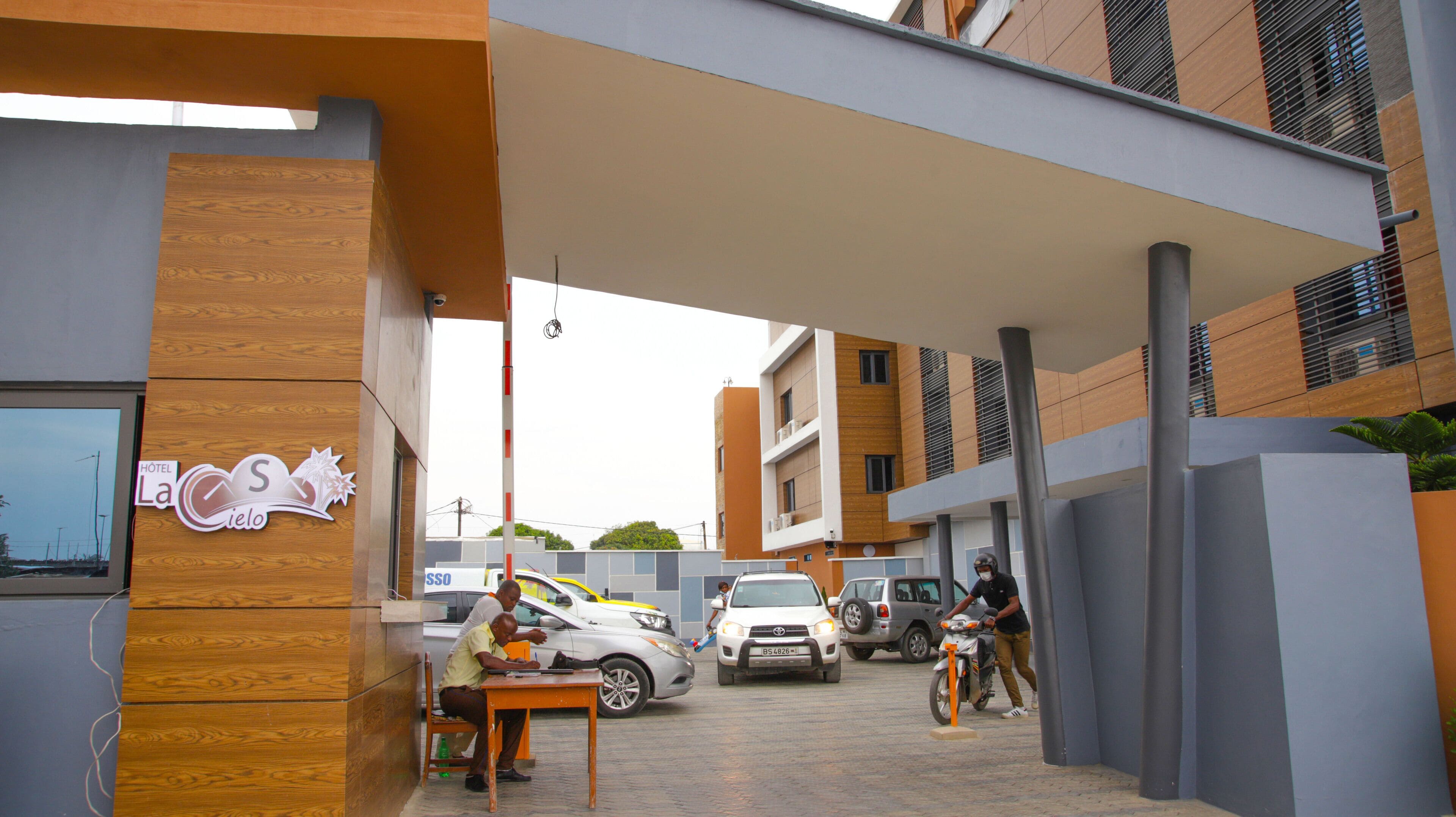 Entrada de hotel com um letreiro, área de estacionamento coberta e uma pessoa numa motocicleta.