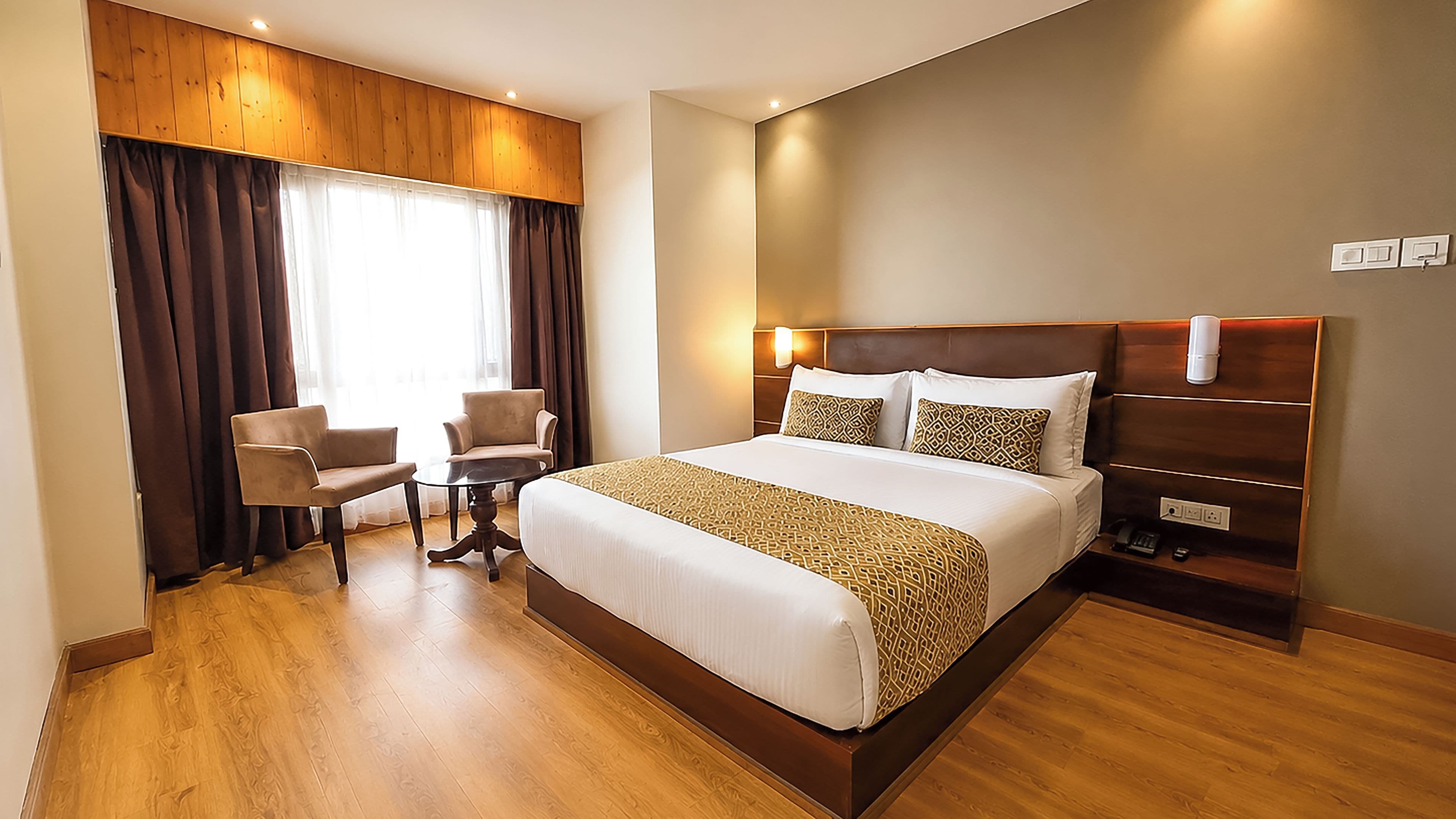 Quarto de hotel com uma cama grande, uma escrivaninha, duas cadeiras, chão de madeira e uma janela com cortinas.