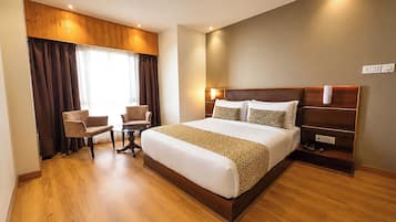 Quarto de hotel com uma cama grande, uma escrivaninha, duas cadeiras, chão de madeira e uma janela com cortinas.