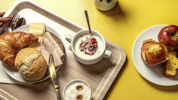 Uma bandeja com croissants, um sanduíche, uma tigela de iogurte com fruta, um muffin e uma maçã sobre uma mesa amarela.