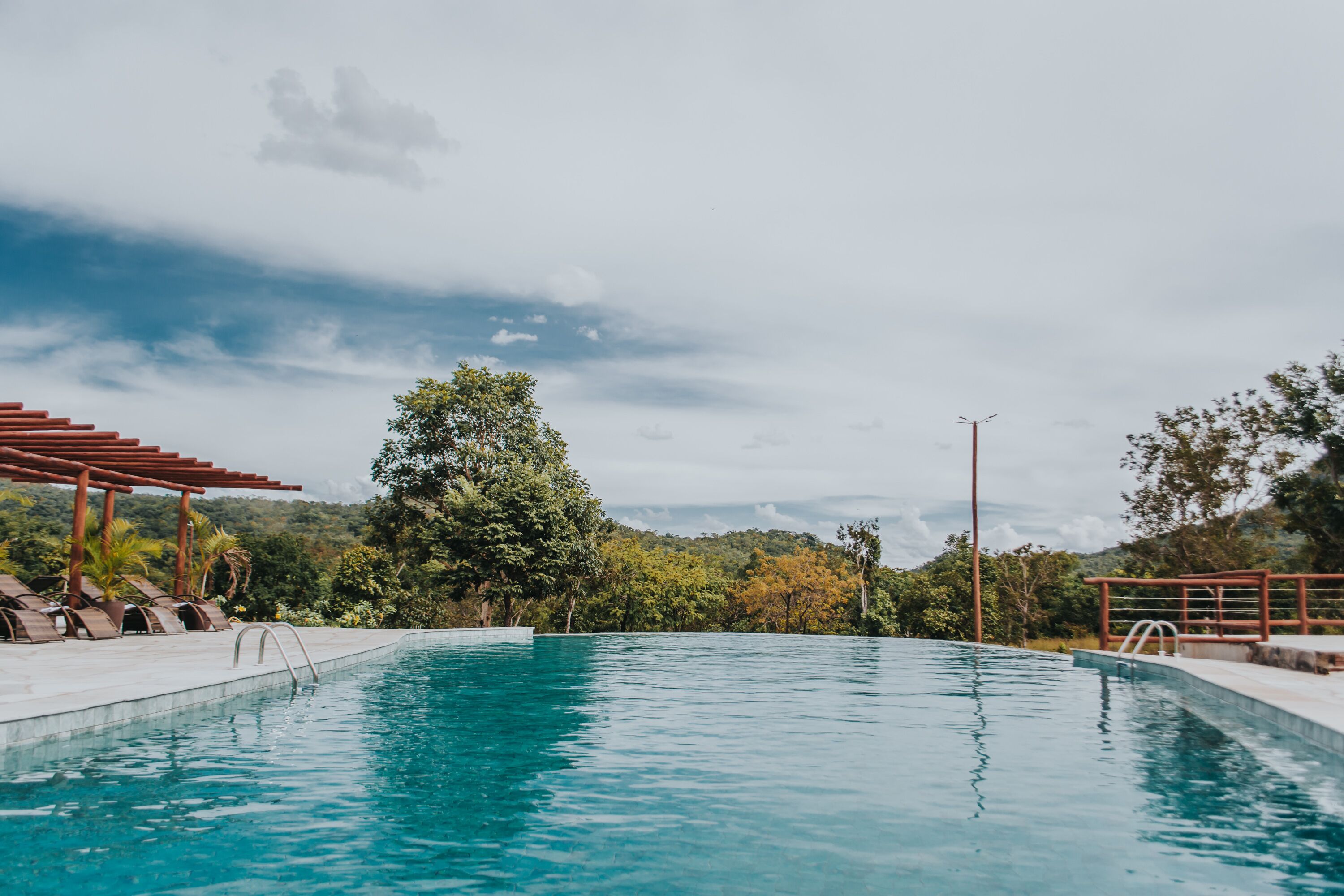 Uma piscina azul cristalina com um pérgola de madeira e espreguiçadeiras, rodeada por vegetação exuberante e árvores.
