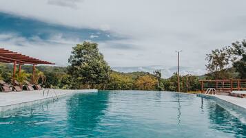 Uma piscina azul cristalina com um pérgola de madeira e espreguiçadeiras, rodeada por vegetação exuberante e árvores.