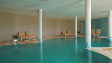 Uma grande piscina interior com água azul cristalina, rodeada por colunas brancas e espreguiçadeiras com almofadas laranja.