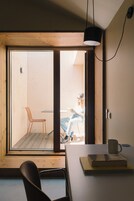 Quarto moderno com porta de vidro deslizante que dá para uma área externa, uma cadeira de madeira, uma mesa com um livro e uma caneca, e um candeeiro pendente.