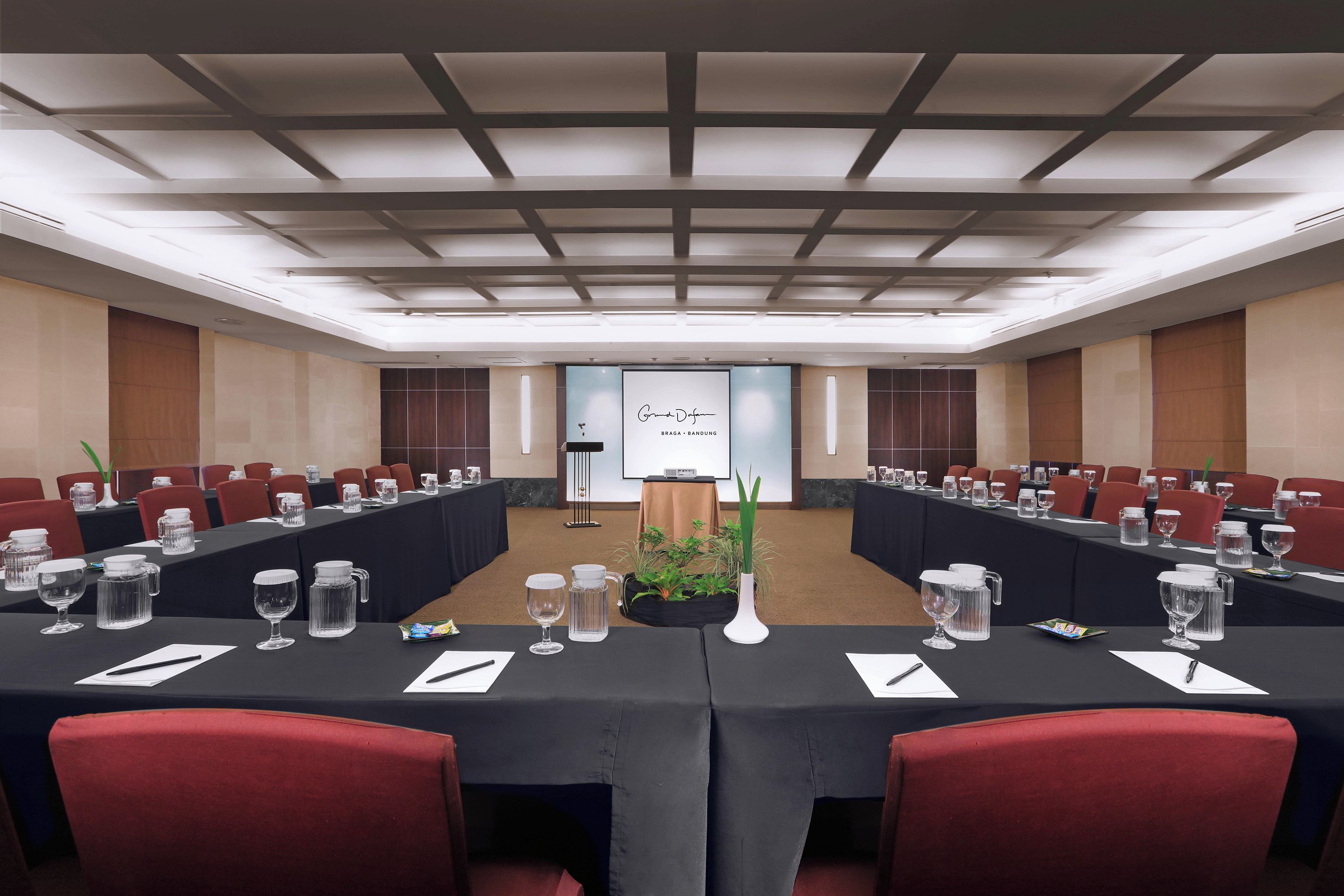 Uma sala de conferências com uma mesa comprida, cadeiras vermelhas e uma tela de apresentação.