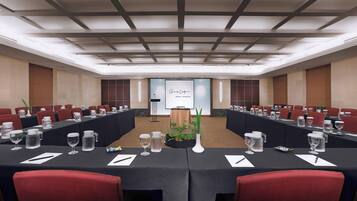 Uma sala de conferências com uma mesa comprida, cadeiras vermelhas e uma tela de apresentação.
