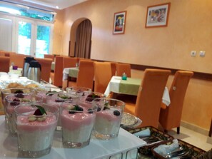Uma área de refeições com mesas postas para uma refeição, com um buffet de sobremesas e diversas opções de frutas e oleaginosas.