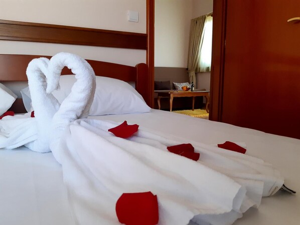 Quarto de hotel com cama, cabeceira de madeira, mesa de cabeceira com abajur e janela com cortinas.