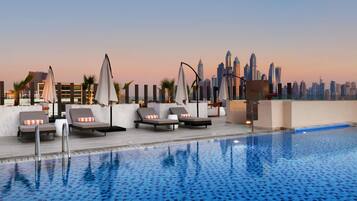Piscina no terraço com espreguiçadeiras e guarda-sóis, com vista para o horizonte da cidade ao pôr do sol.