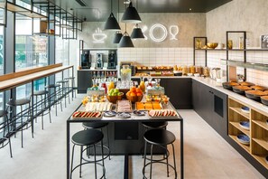 Um café moderno com uma mesa comunitária grande, banquetas no balcão e diversas opções de comida e bebida.